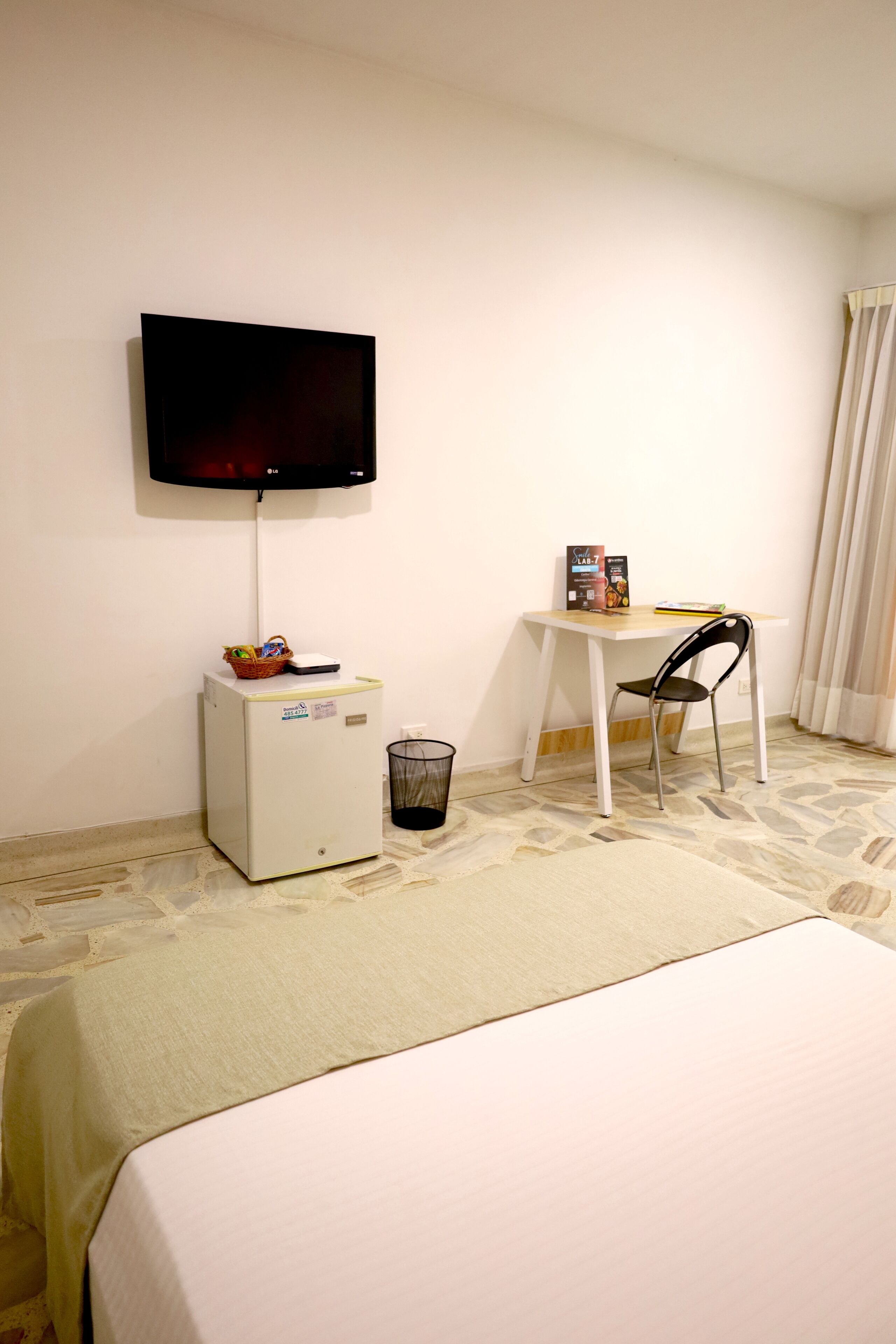 Quarto de hotel com cama, escrivaninha, cadeira, televisão, mini-geladeira e lixeira.