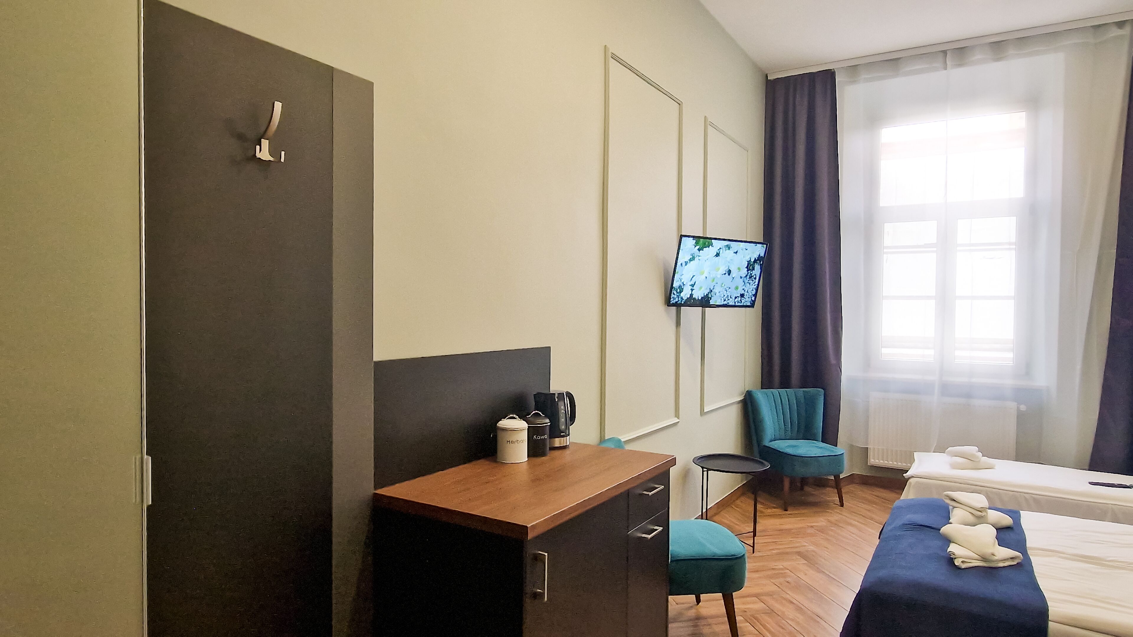 Quarto de hotel com uma escrivaninha de madeira, uma cadeira, televisão presa na parede, cama com colcha azul e uma janela com cortinas.