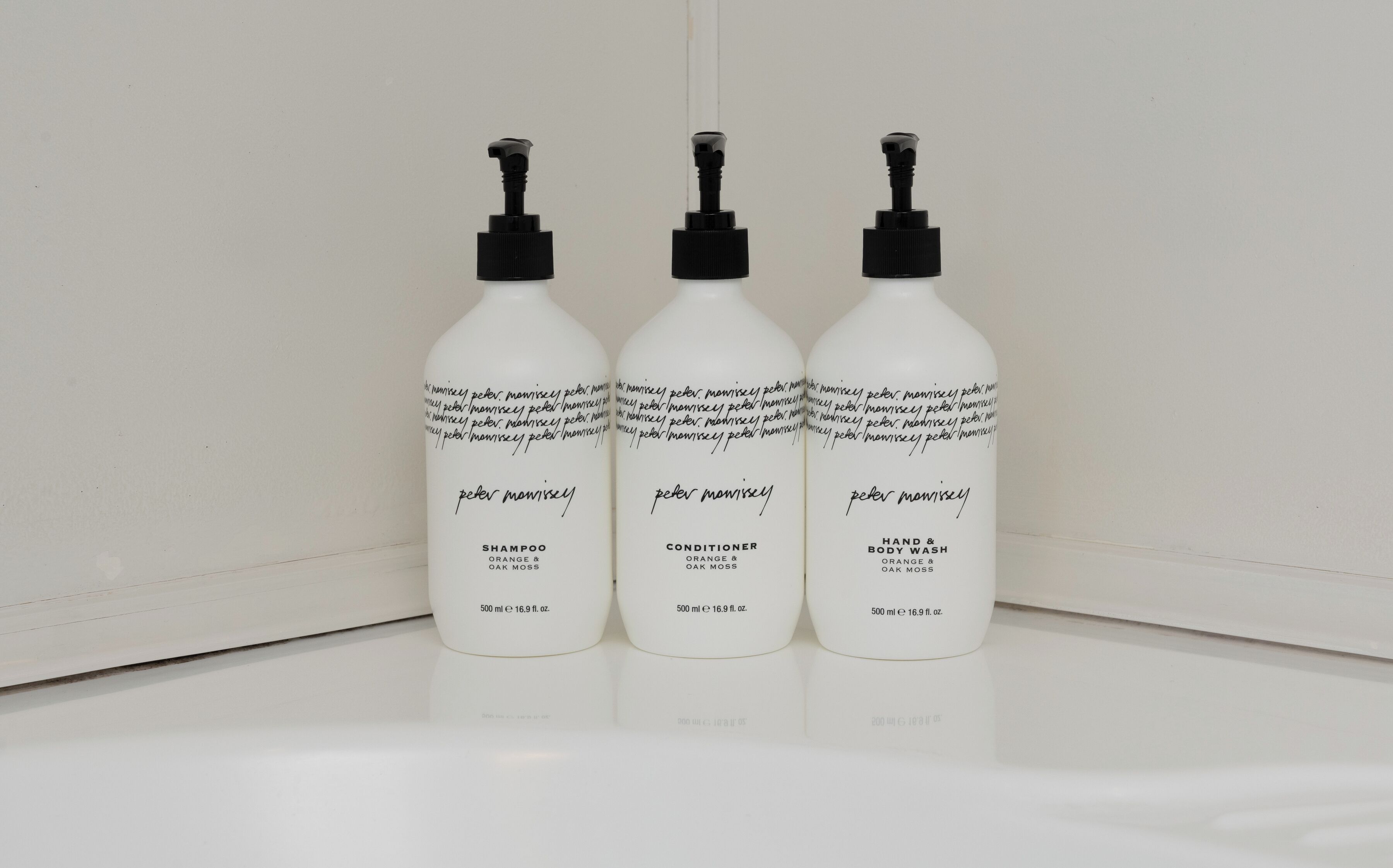 Três frascos de produtos Peter Monissey: shampoo de laranja, condicionador e sabonete líquido para mãos e corpo, cada um com o nome do produto e volume indicado.