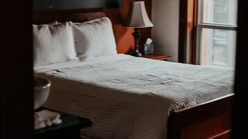Cama bem arrumada com edredom branco e almofadas, mesa de cabeceira com abajur e um pôster na parede.