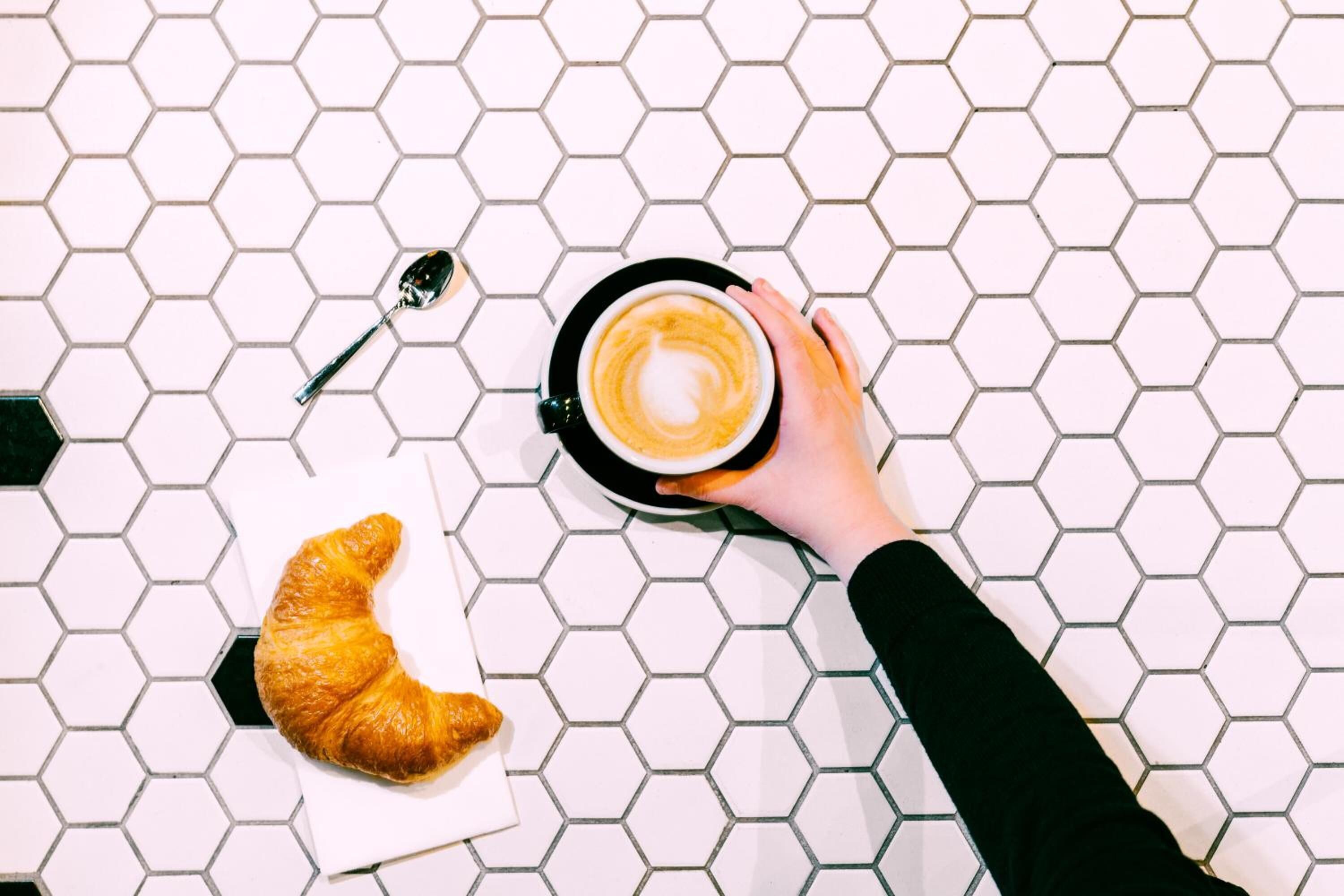 Vista de cima de um chão com ladrilhos hexagonais, com um croissant e uma xícara de café sobre uma superfície branca.