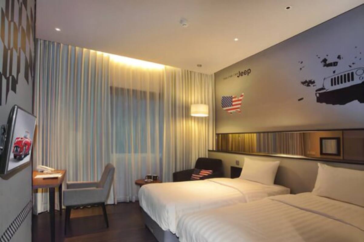 Quarto de hotel com cama, escrivaninha, cadeira e parede com o logotipo da Jeep e uma bandeira americana.