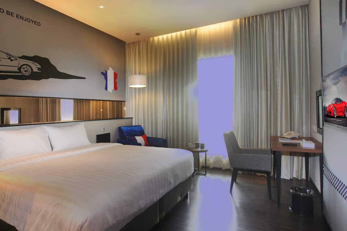 Quarto de hotel com cama grande, escrivaninha com cadeira, televisão e parede com imagem de carro.