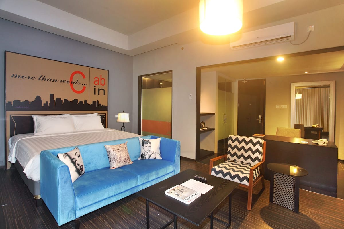Quarto de hotel com uma cama, um sofá azul, uma cadeira de madeira, uma mesinha e um espelho.