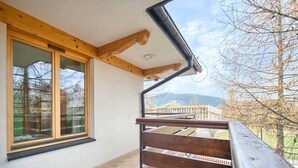 Uma varanda com um banco de madeira, uma janela com vista para edifícios e árvores, e um corrimão de metal.