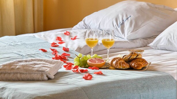 Cama bem arrumada com travesseiro branco, dois copos de suco de laranja, uma tigela de uvas, meia toranja e um prato de croissants.