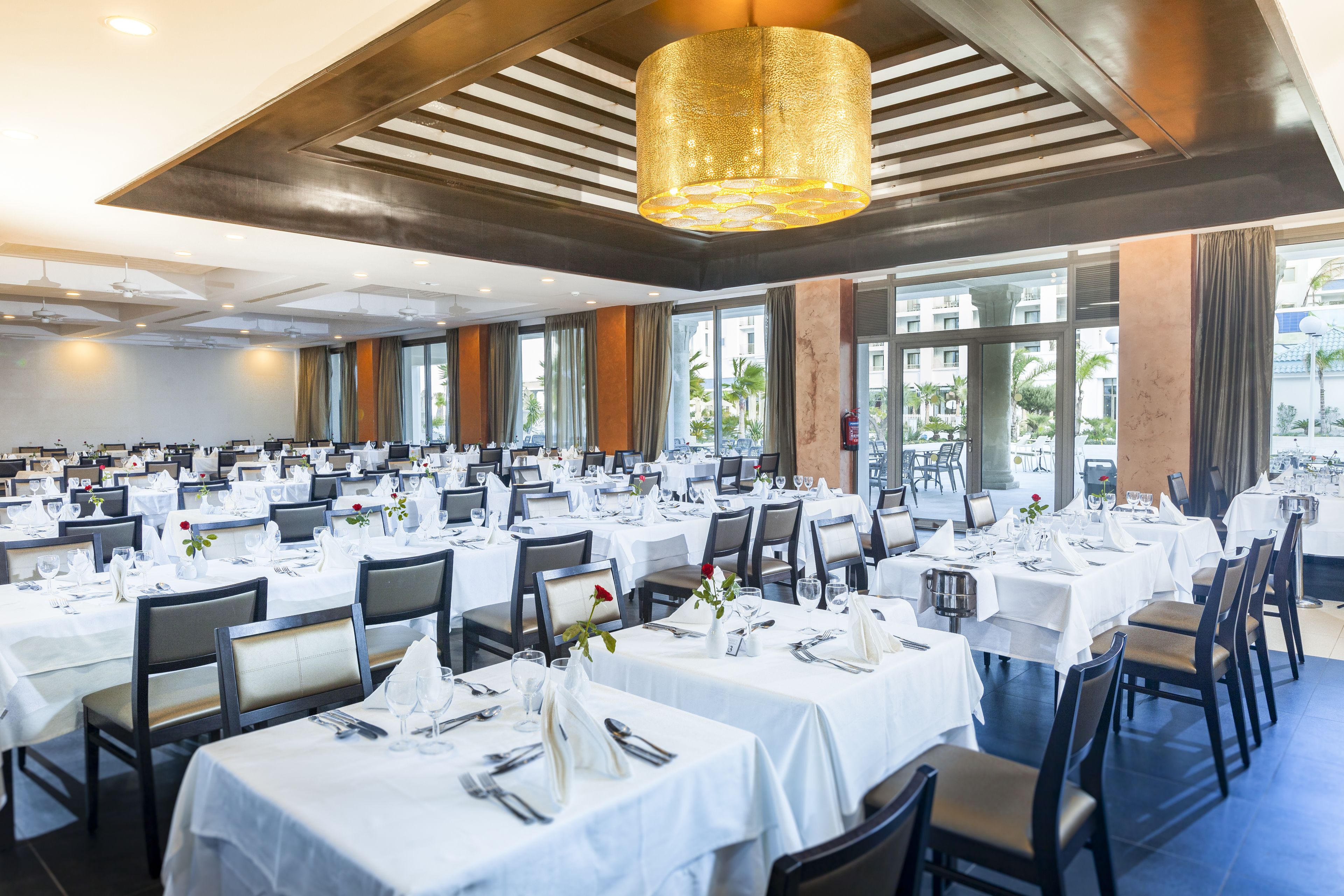 Ampla sala de jantar com várias mesas preparadas para serviço, destacando um design moderno no teto e grandes janelas com vista para o exterior.