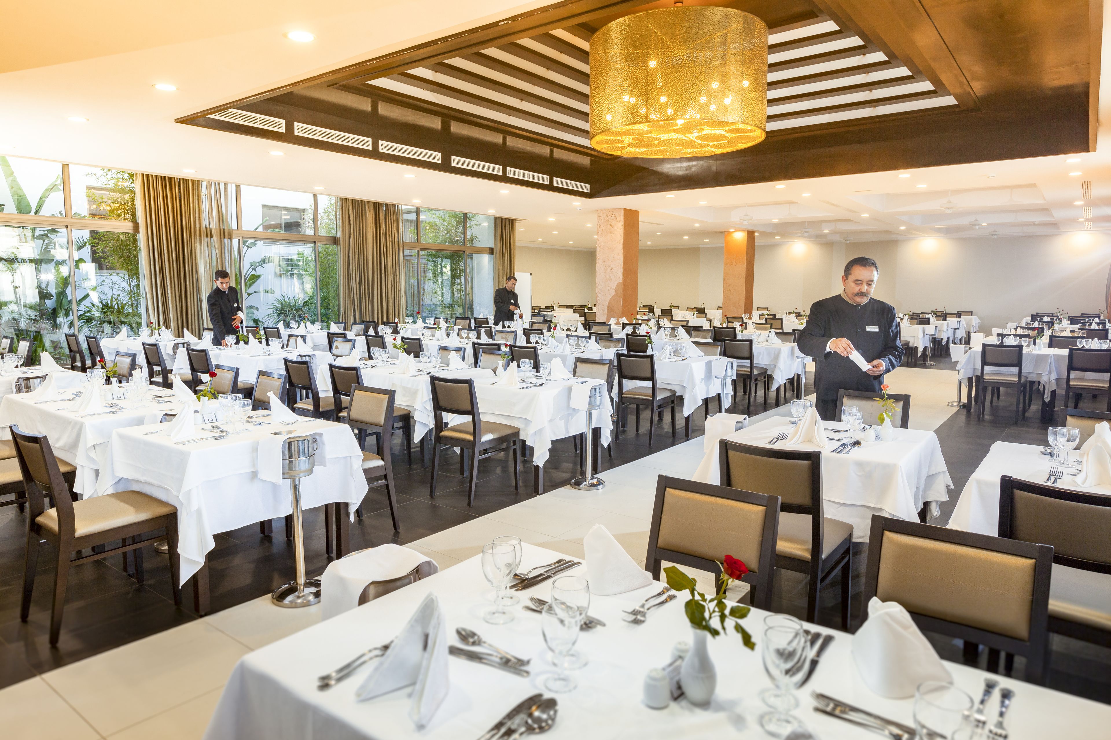 Um amplo salão de jantar com várias mesas montadas para um evento formal, com toalhas brancas e talheres.