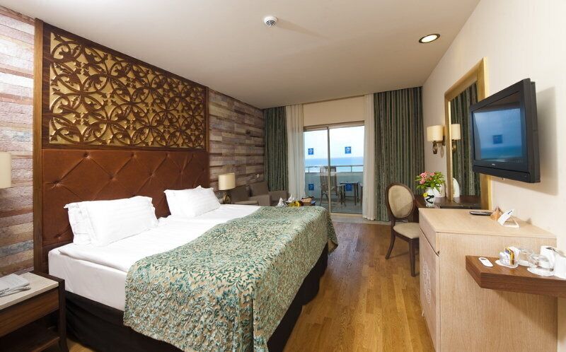 Quarto de hotel com uma cama grande, uma mesa de trabalho com televisão e vista para o mar pela janela.