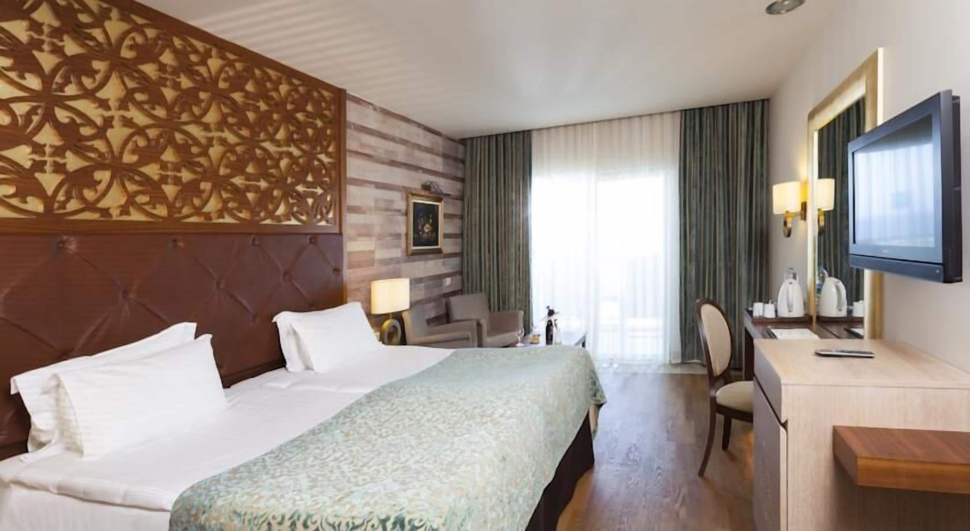Quarto de hotel com uma cama grande, uma escrivaninha com cadeira, televisão e janela com cortinas.