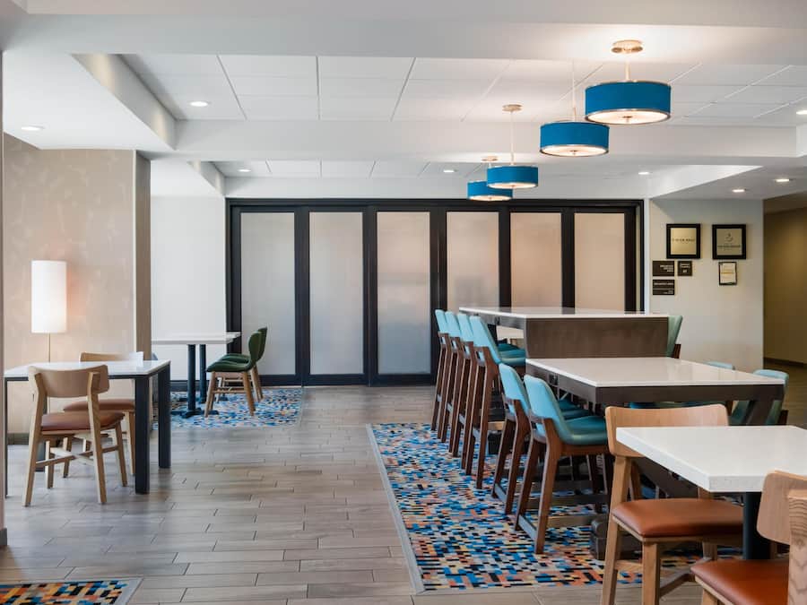 Uma sala de jantar moderna com várias mesas e cadeiras, um tapete estampado e luminárias pendentes azuis.