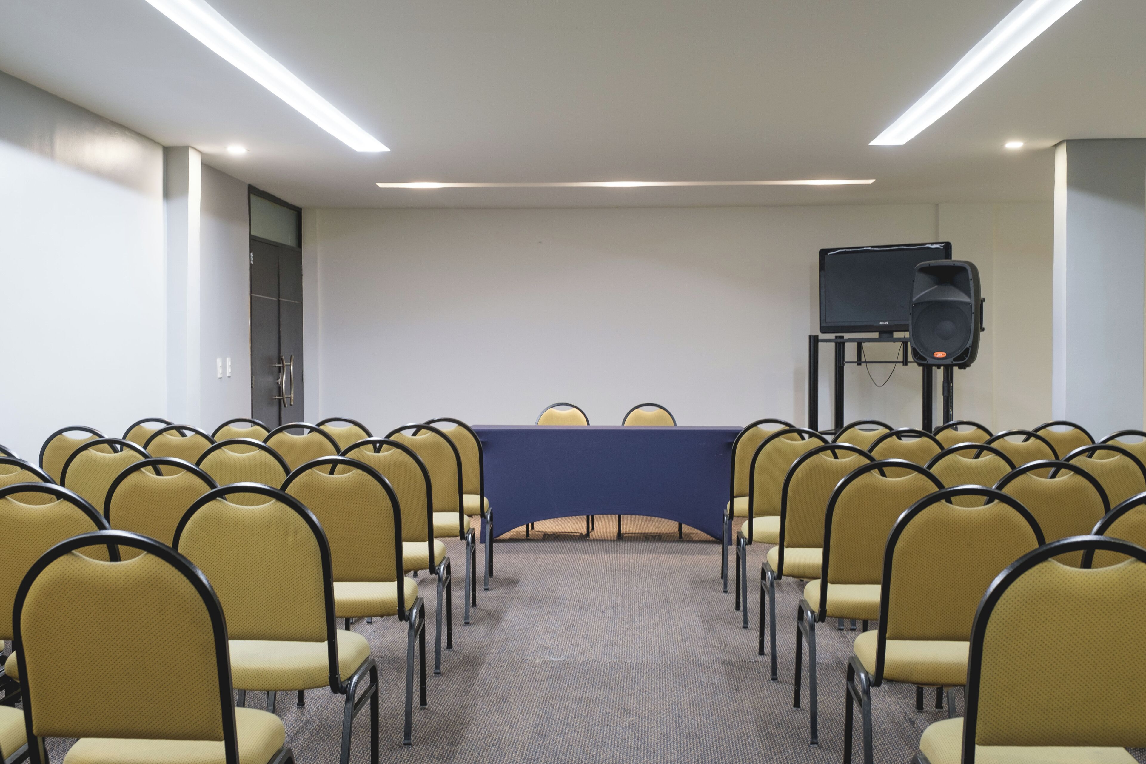 Uma sala de conferências com uma mesa comprida, cadeiras amarelas, toalha de mesa azul, um alto-falante em um suporte e uma porta.