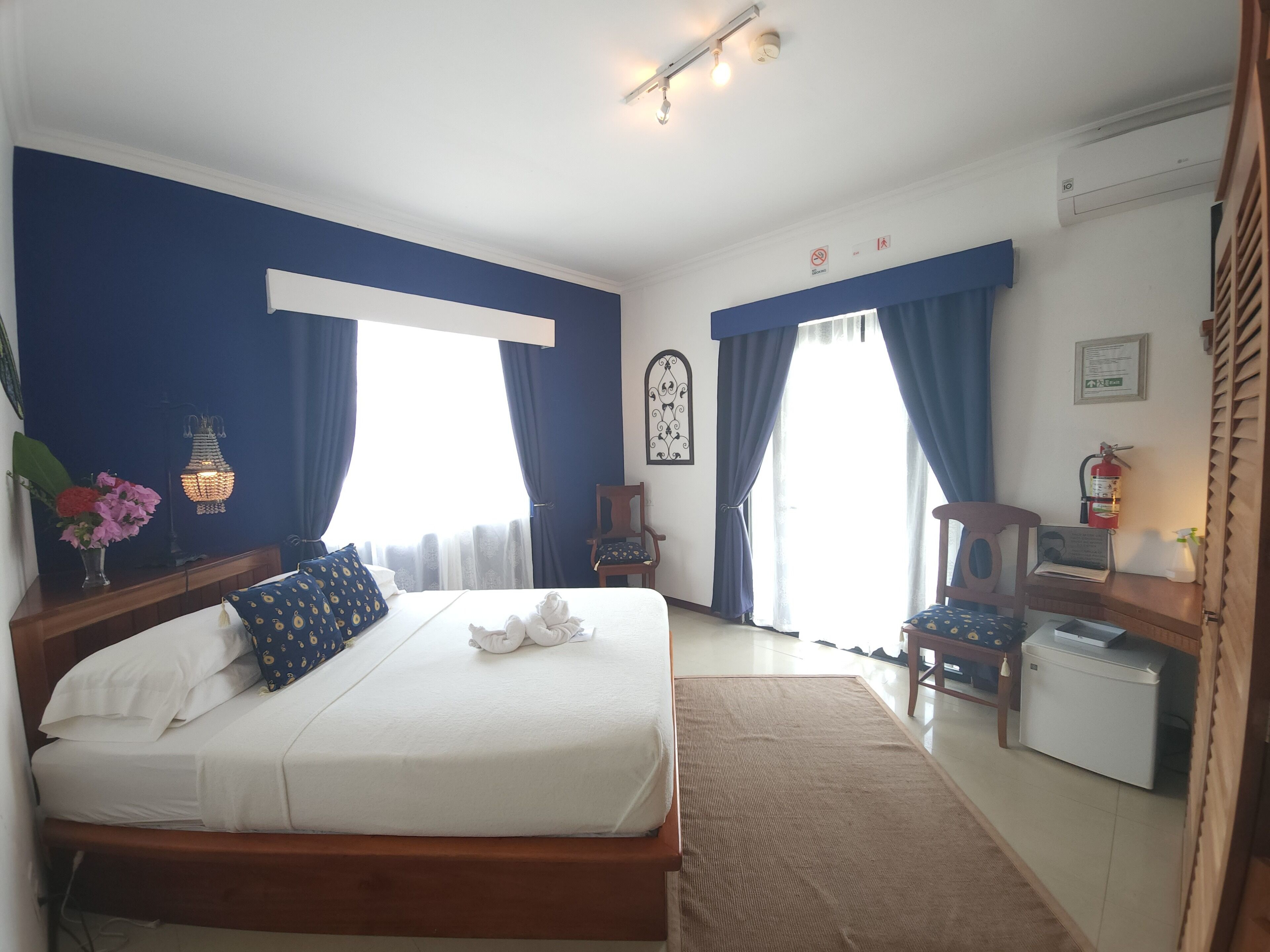 Quarto de hotel com cama, escrivaninha, cadeira, pequeno frigorífico e janela com cortinas.