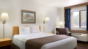 Quarto de hotel com cama, duas luminárias de cabeceira, uma cadeira, janela com cortinas e um quadro na parede.
