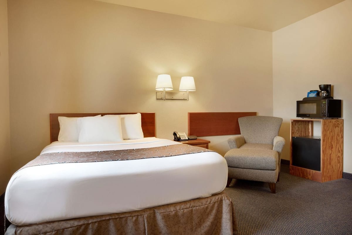 Quarto de hotel com uma cama grande, duas luminárias de cabeceira, uma cadeira, uma escrivaninha com telefone e um micro-ondas em um armário de madeira.