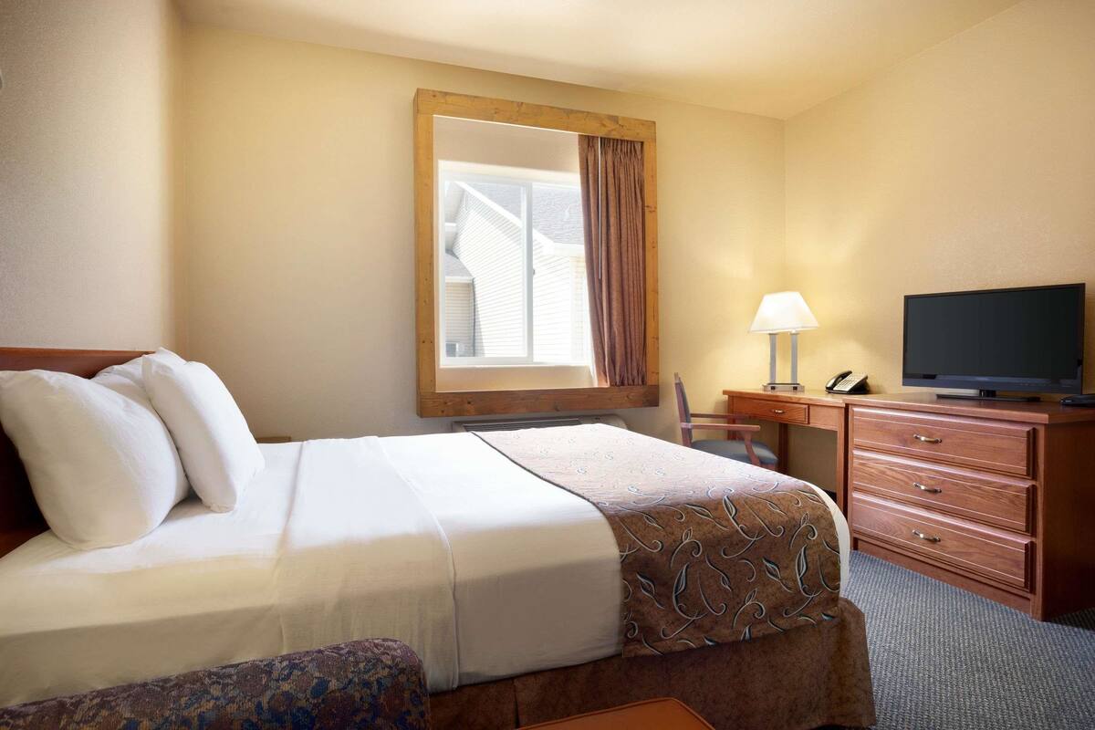 Quarto de hotel com cama, escrivaninha com luminária, televisão e uma janela grande com cortinas.