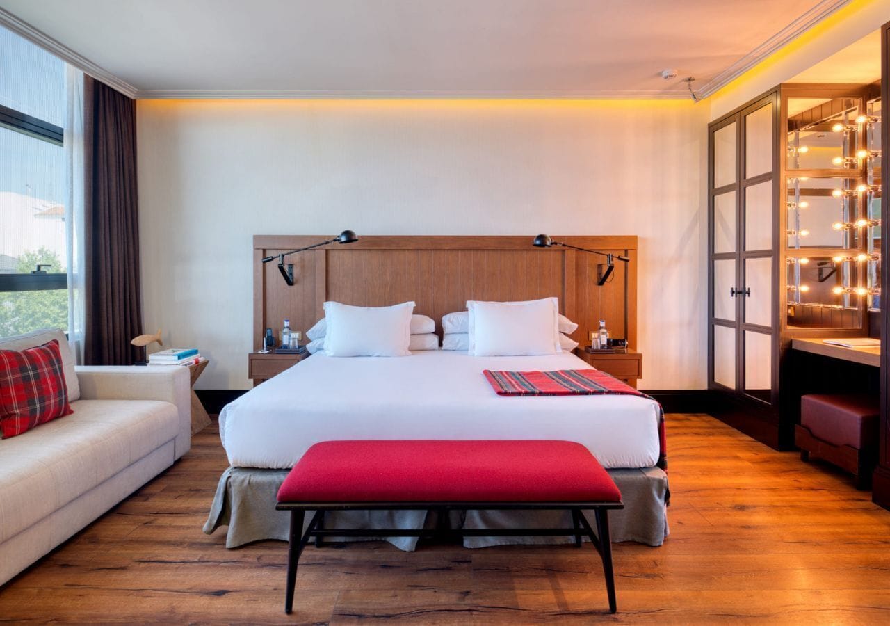 Quarto de hotel moderno com cama grande, mesa de cabeceira, um banco vermelho e um espelho com iluminação.