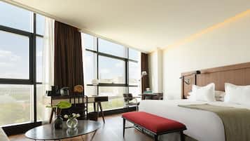 Quarto de hotel com uma grande janela, uma cama com roupa branca, uma escrivaninha com cadeira, uma pequena mesa com um vaso e um banco vermelho.