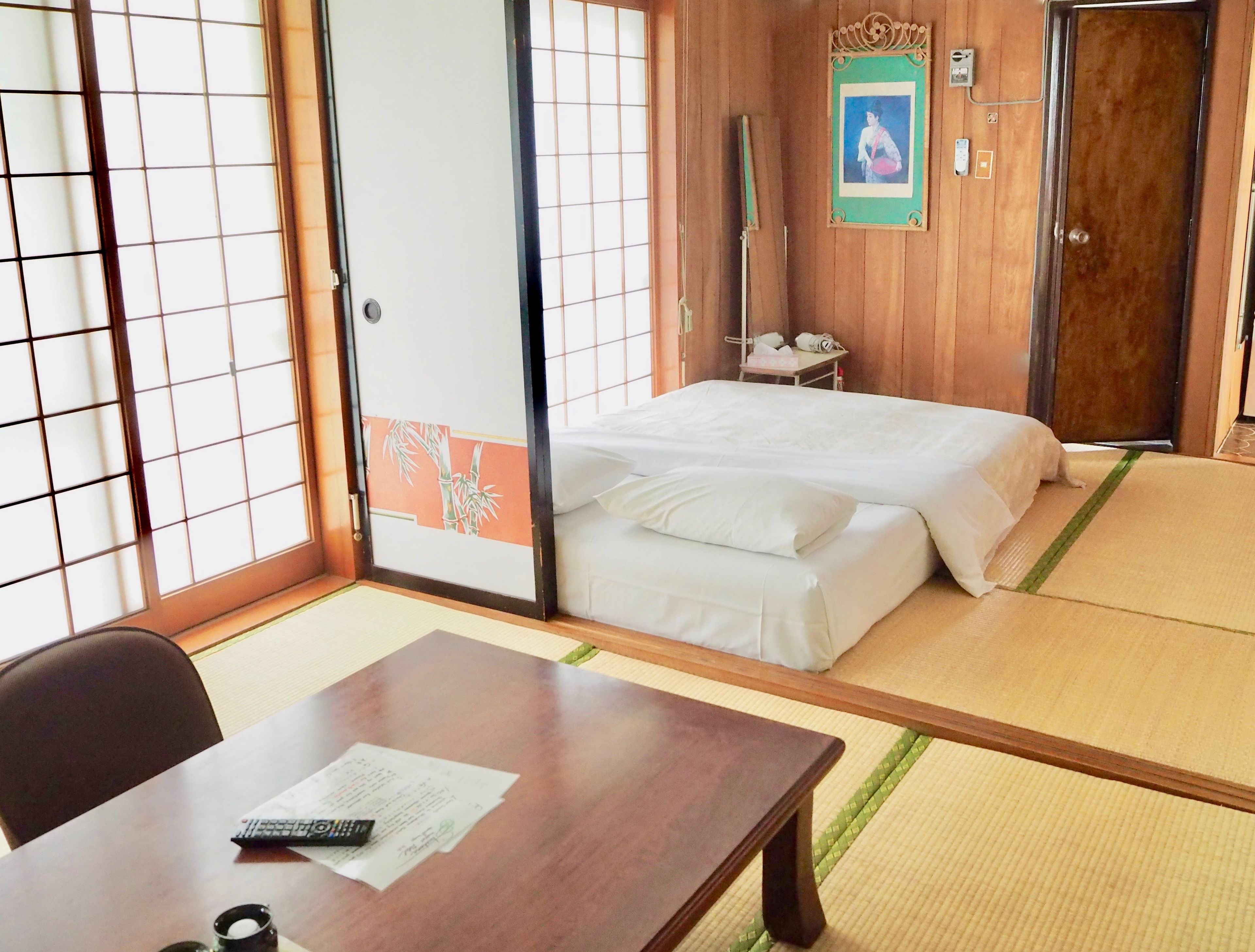 Quarto japonês tradicional com tatames, uma cama com roupa de cama branca, uma escrivaninha com um controle remoto e um quadro na parede.
