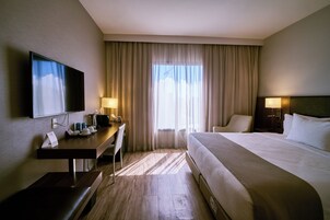 Quarto de hotel com cama, escrivaninha, cadeira, televisão e cortinas.