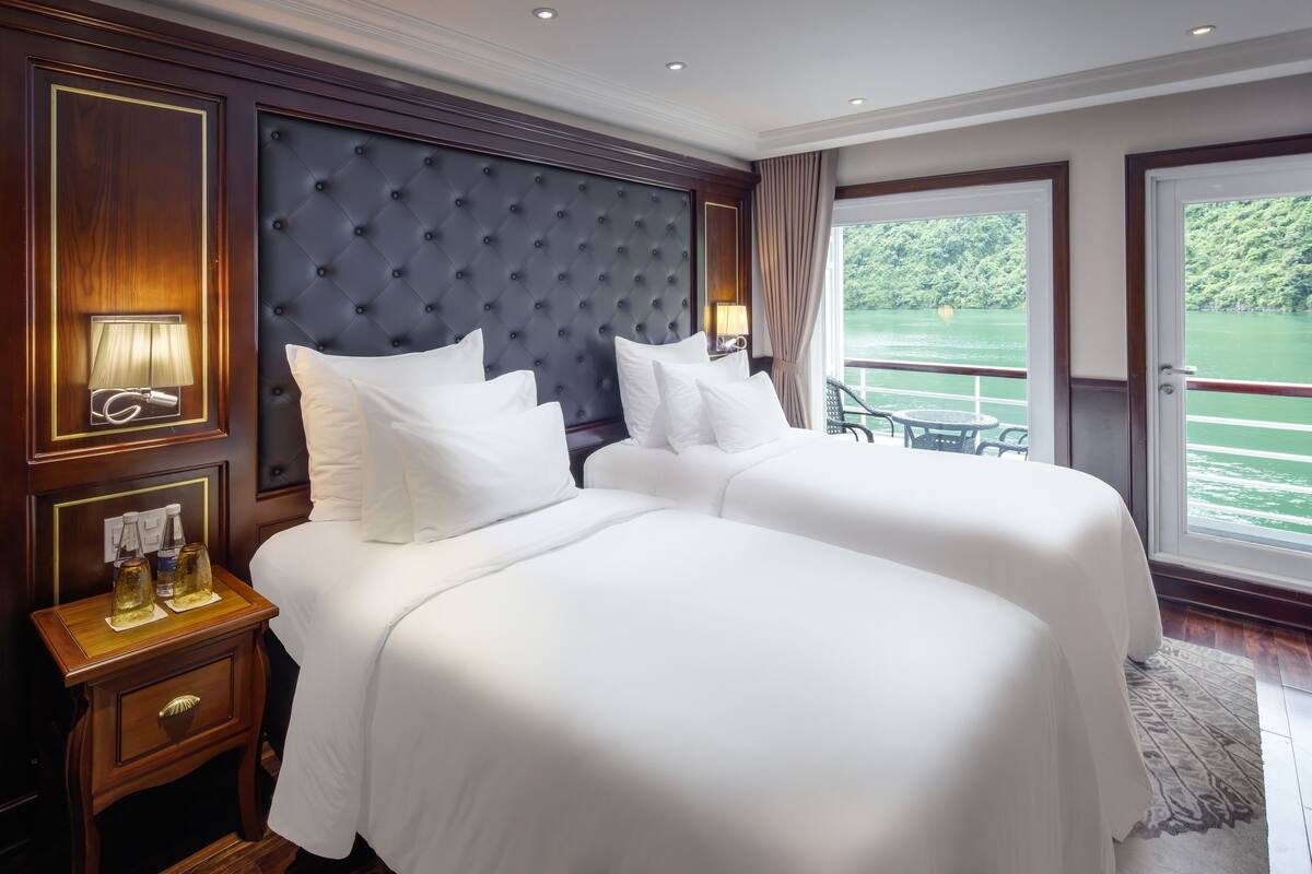 Quarto de hotel com duas camas, uma cabeceira de madeira, uma mesa de cabeceira com abajur e vista para uma paisagem verde pela janela.