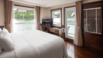 Quarto de hotel com uma cama grande, uma mesa, uma cadeira e vista para um lago através de janelas amplas.