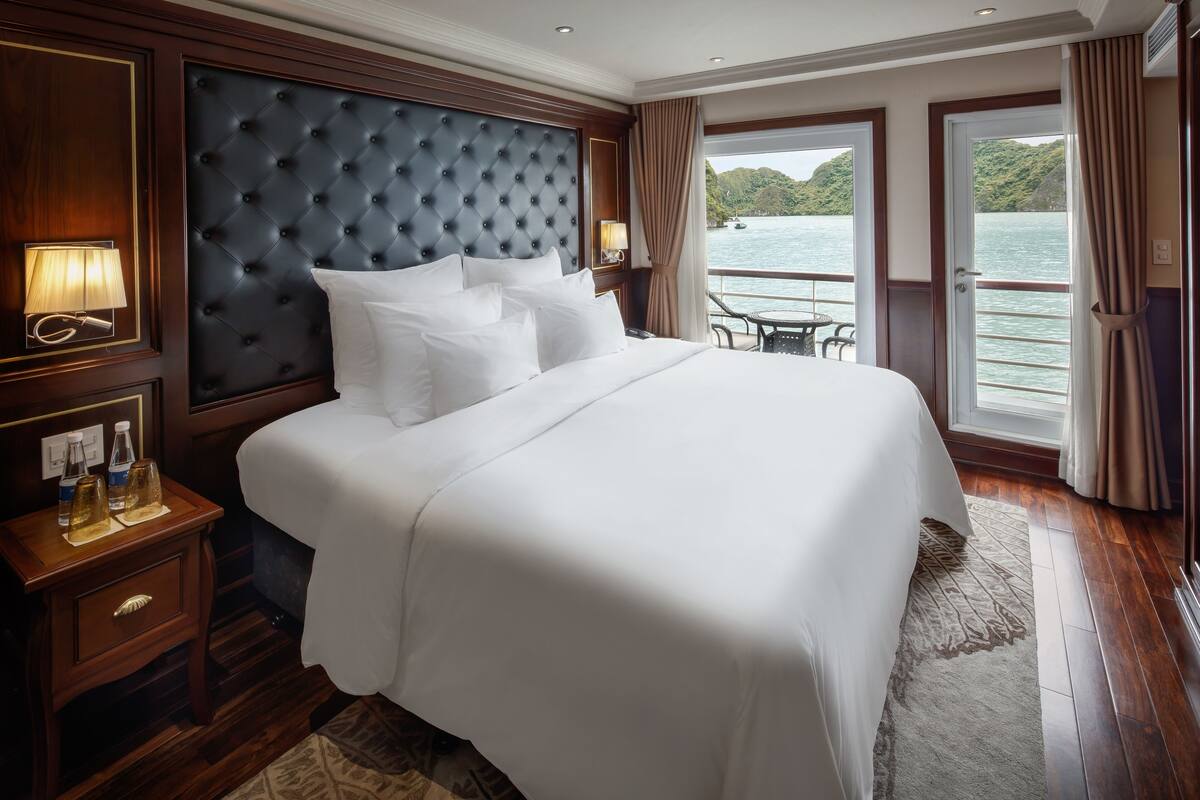 Uma cama grande com cabeceira estofada, roupa de cama branca, uma mesa de cabeceira de madeira com abajur e vista para a água através de uma porta de vidro.