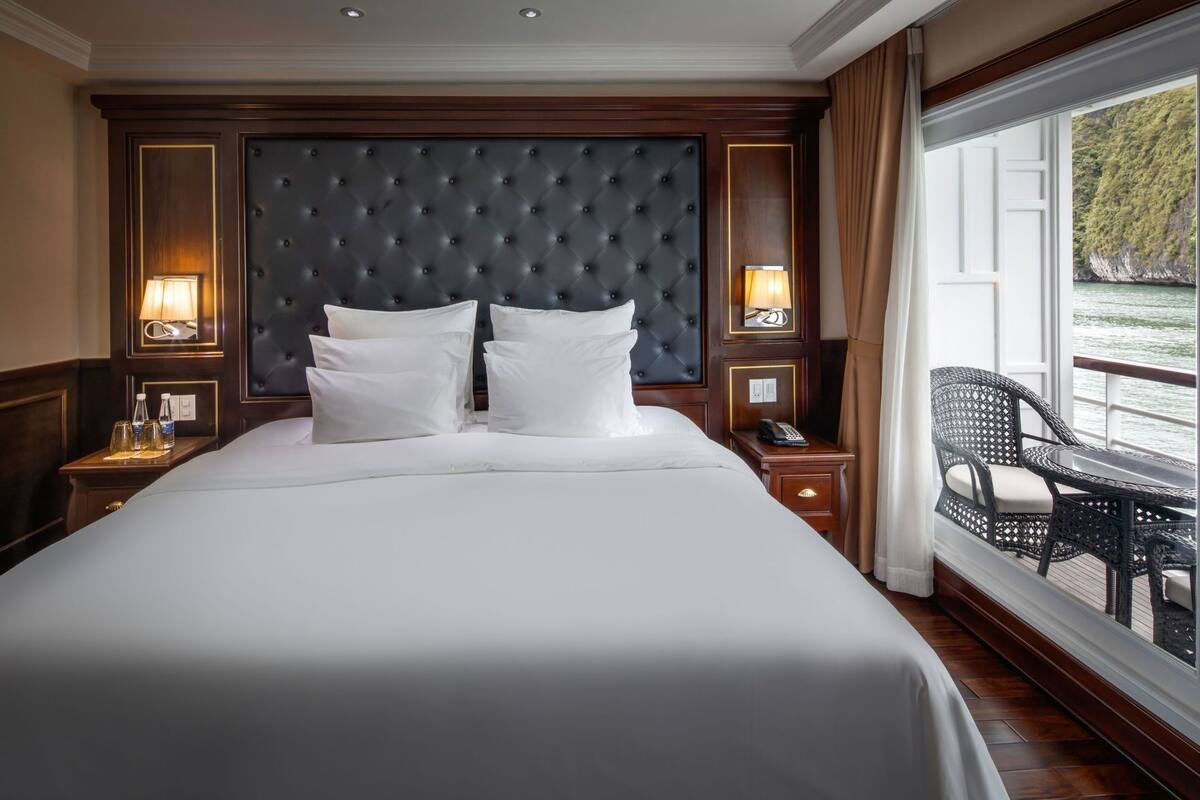 Quarto de hotel com uma cama grande, duas mesas de cabeceira, uma cabeceira escura com efeito acolchoado e vista para a água através de uma janela ampla.