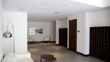 Um lobby moderno de hotel com um sofá branco, uma mesa pequena, iluminação embutida no teto, uma janela grande e uma porta com padrão quadriculado.