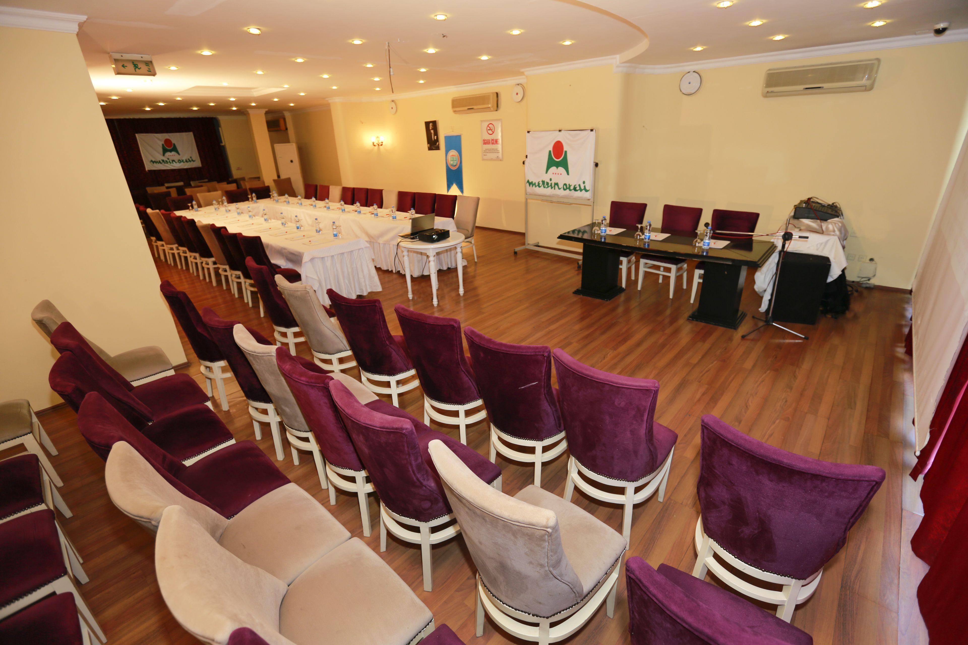 Uma sala de conferências com fileiras de cadeiras roxas, uma mesa comprida com toalha branca e um palco com um banner.