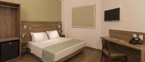 Quarto de hotel com cama, escrivaninha, cadeira, televisão e um pequeno frigorífico.