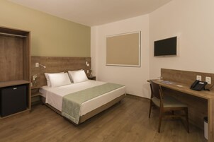 Quarto de hotel com cama, escrivaninha, cadeira, televisão e um pequeno frigorífico.