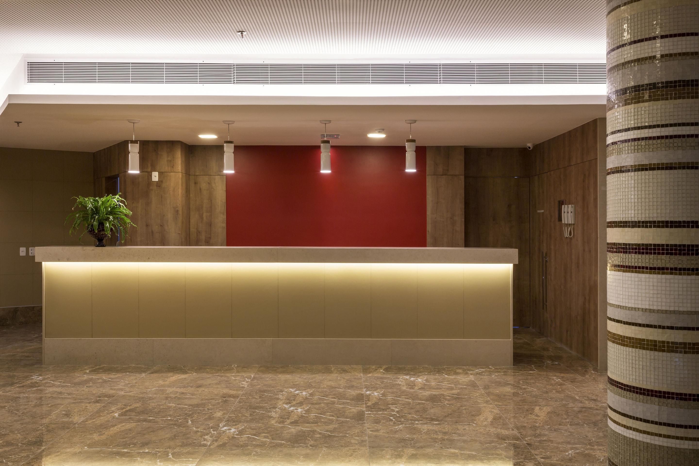 Uma área de recepção com piso de mármore, uma parede com destaque em vermelho e um balcão de atendimento com iluminação suspensa.