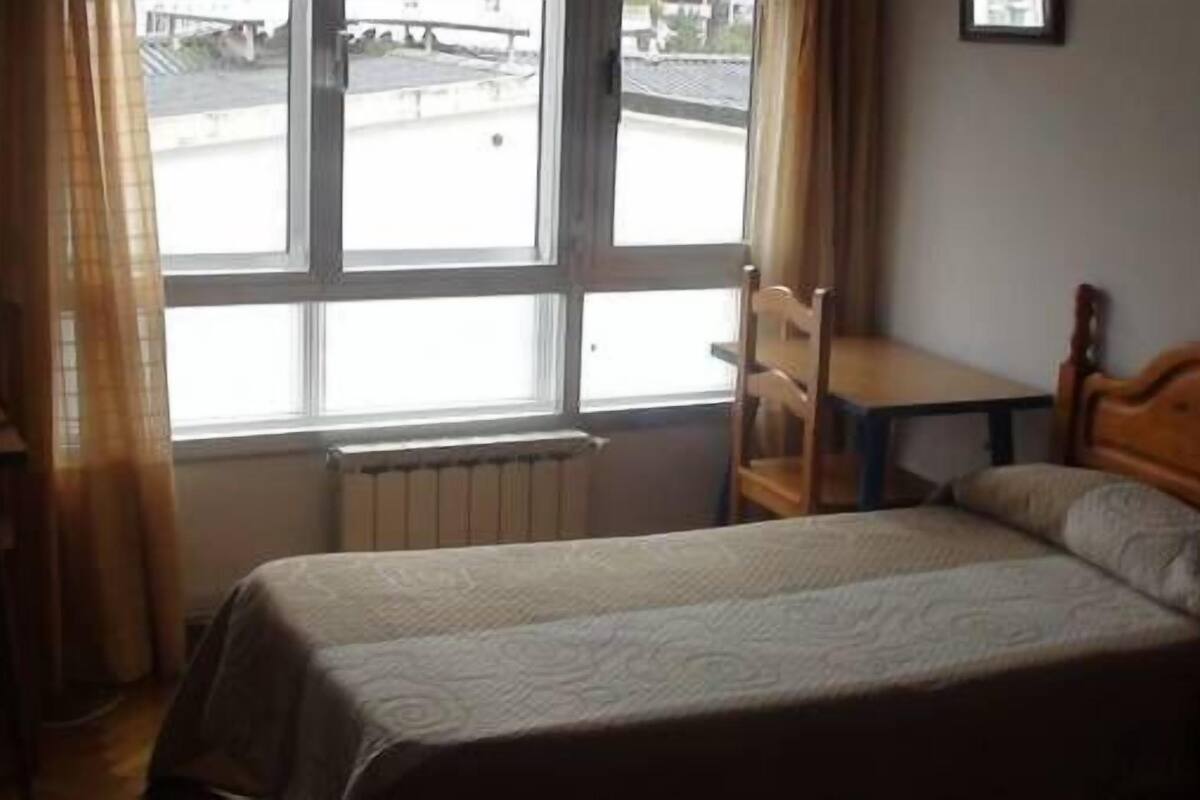 Quarto com cama, mesa pequena, cadeira e janela com vista para uma ponte.