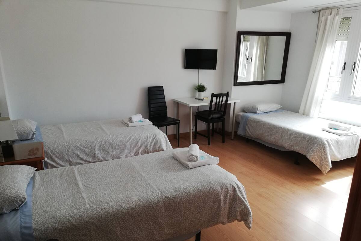 Quarto de hotel com duas camas, uma escrivaninha, uma cadeira, uma televisão e uma janela com cortinas.