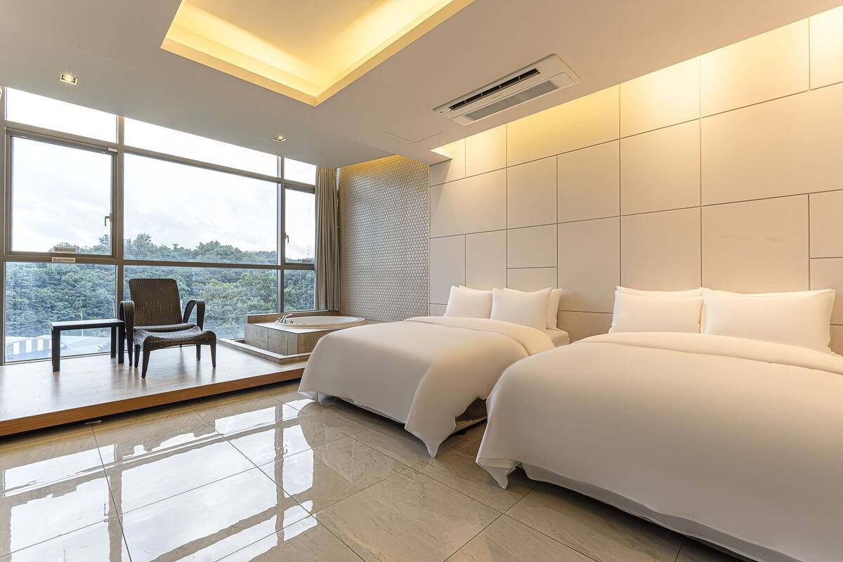 Quarto de hotel moderno com duas camas, uma janela ampla e uma vista, uma mesinha e uma cadeira.