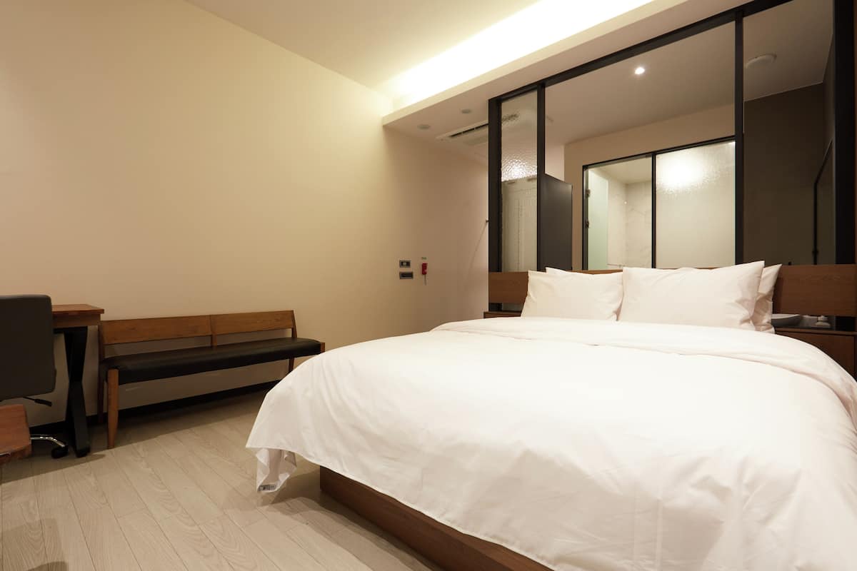 Quarto de hotel moderno com uma cama grande, um banco de madeira, uma escrivaninha e um espelho amplo.
