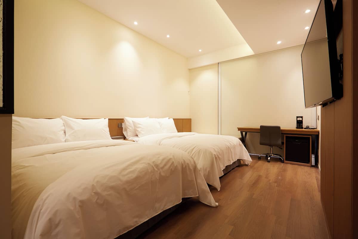 Quarto de hotel com duas camas, uma escrivaninha, uma cadeira e uma televisão fixada na parede.