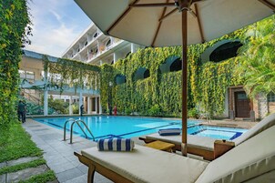 Piscina com espreguiçadeiras e área coberta, cercada por vegetação e um edifício com várias sacadas.