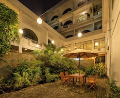 Um pátio com mesa e cadeiras sob guarda-sol, cercado por vegetação exuberante e um edifício com várias sacadas.
