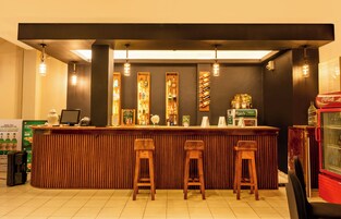 Uma área de bar com balcão de madeira, três banquetas e prateleiras com garrafas.