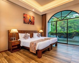 Quarto de hotel com uma cama grande, duas mesas de cabeceira, um criado-mudo, um quadro e uma janela ampla com vista para o verde.