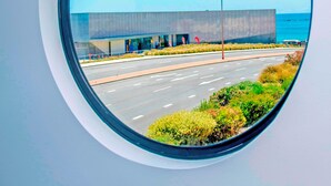 Vista através de uma janela redonda de um edifício moderno, com uma estrada à frente e um corpo d'água ao fundo.