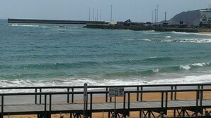 Vista costeira com um cais que se estende para o mar, uma praia de areia e um afloramento rochoso ao longe.