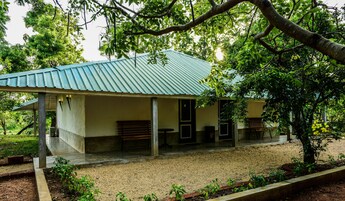 Uma área de estar coberta ao ar livre, com bancos e uma mesa, rodeada por árvores e cascalho.