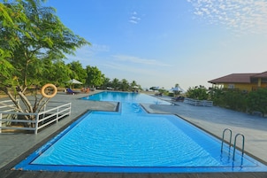 Piscina com água azul cristalina, rodeada por espreguiçadeiras e guarda-sóis, com árvores e um edifício ao fundo.
