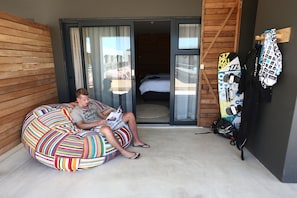 Uma pessoa sentada em um puff colorido lendo um livro em um quarto com porta de vidro deslizante que dá para uma varanda.