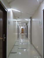 Corredor de hotel com design moderno, com uma porta de vidro à esquerda, uma porta de madeira no final e placas numeradas nas paredes indicando os números dos quartos.