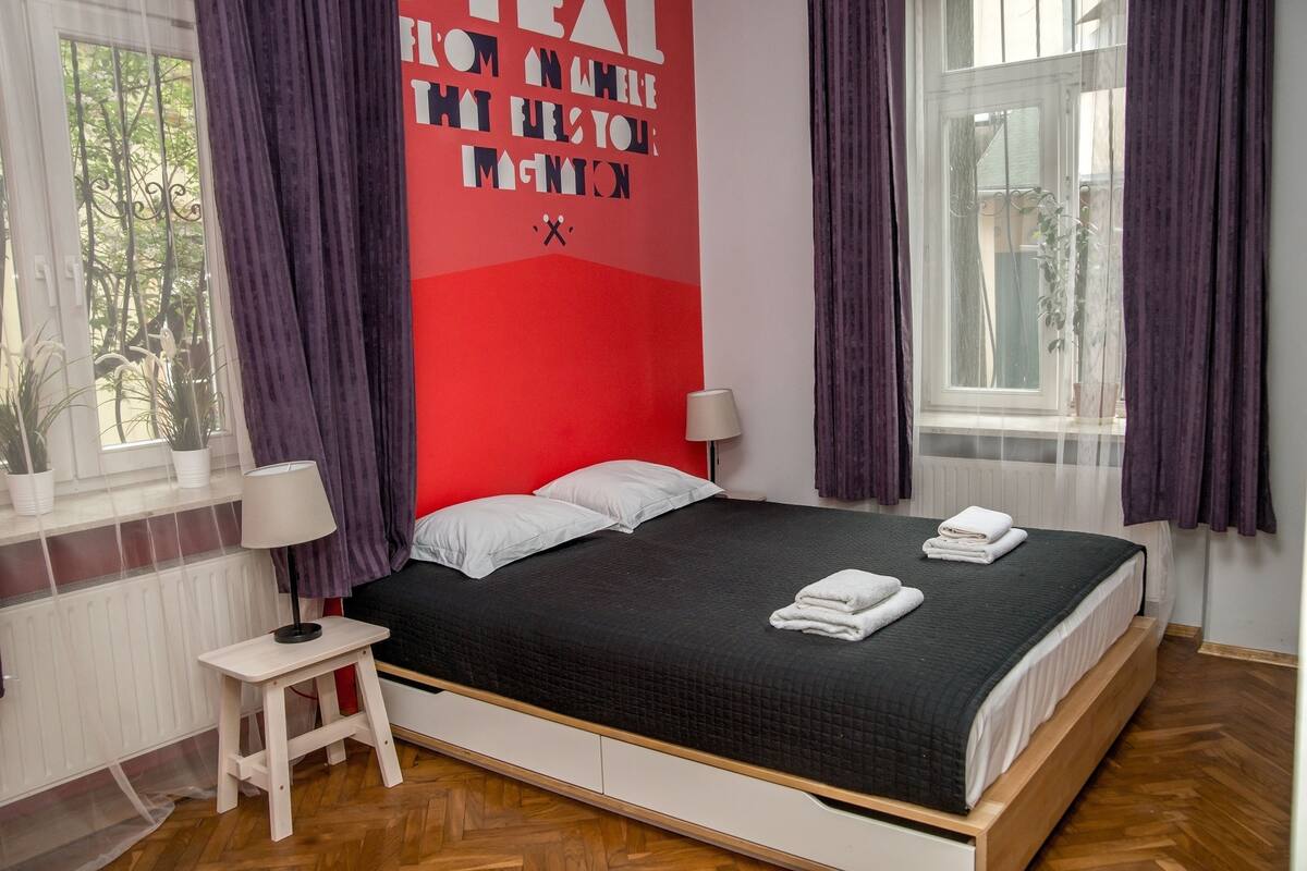 Quarto com cama, mesa de cabeceira, janela com cortinas, radiador e parede com frase motivacional.