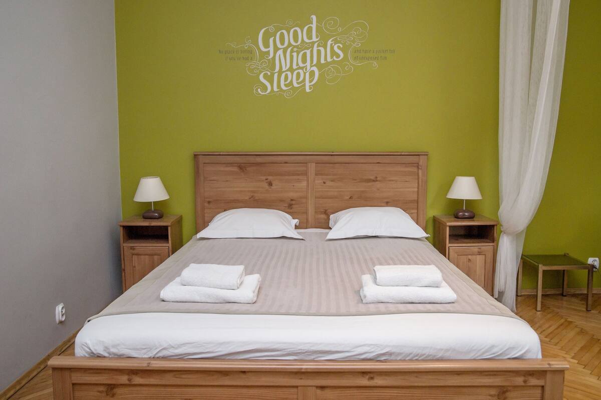 Cama bem arrumada com lençóis e toalhas brancas, uma cabeceira de madeira e duas mesas de cabeceira com abajures.